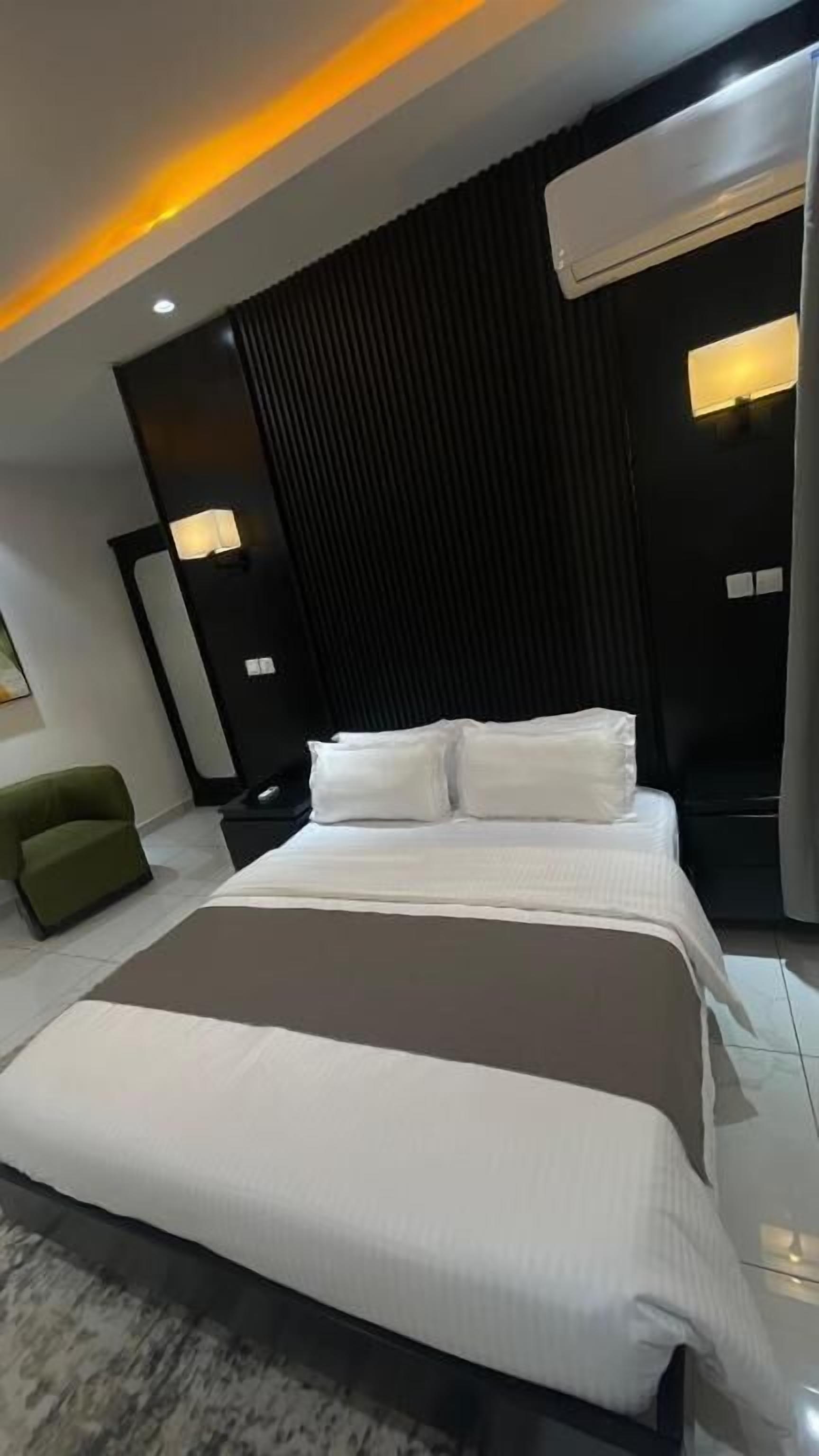 Quarto de hotel moderno com uma cama grande, uma poltrona verde e uma parede de destaque escura.