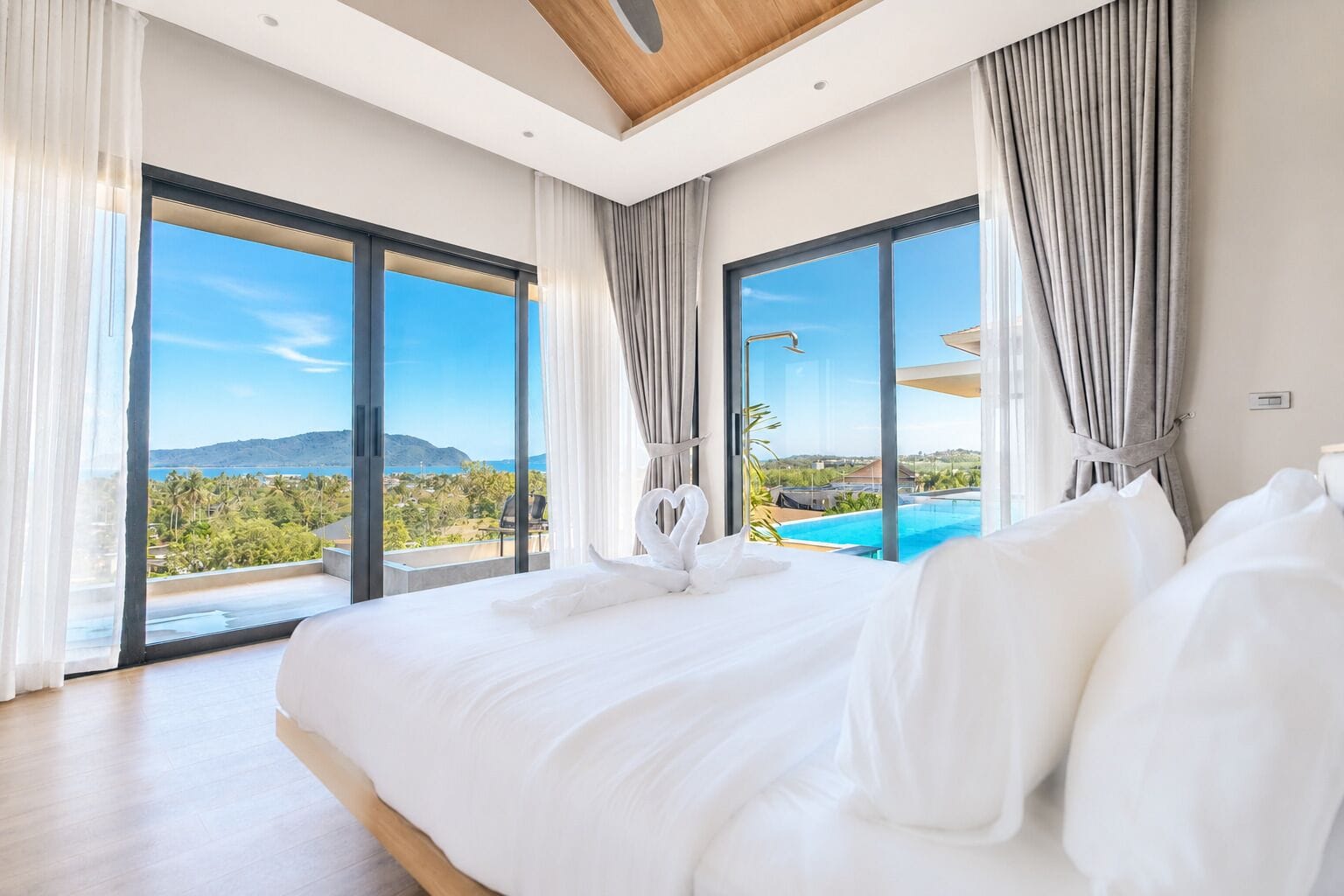 Quarto moderno com cama grande, duas janelas com cortinas, vista para uma piscina e montanhas.