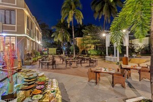 Uma área de refeições ao ar livre com mesas e cadeiras, cercada por palmeiras e um edifício com varanda.