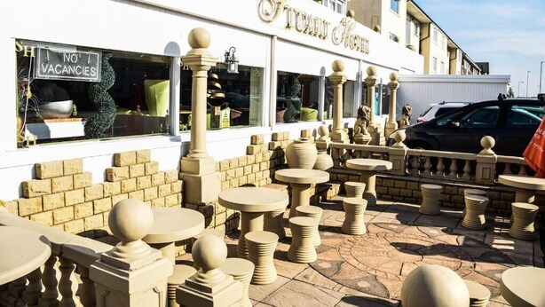 Um edifício com uma placa indicando 'Tudor Furniture', área de estar externa com mesas e cadeiras redondas, e um aviso de 'SEM VAGAS' na janela.