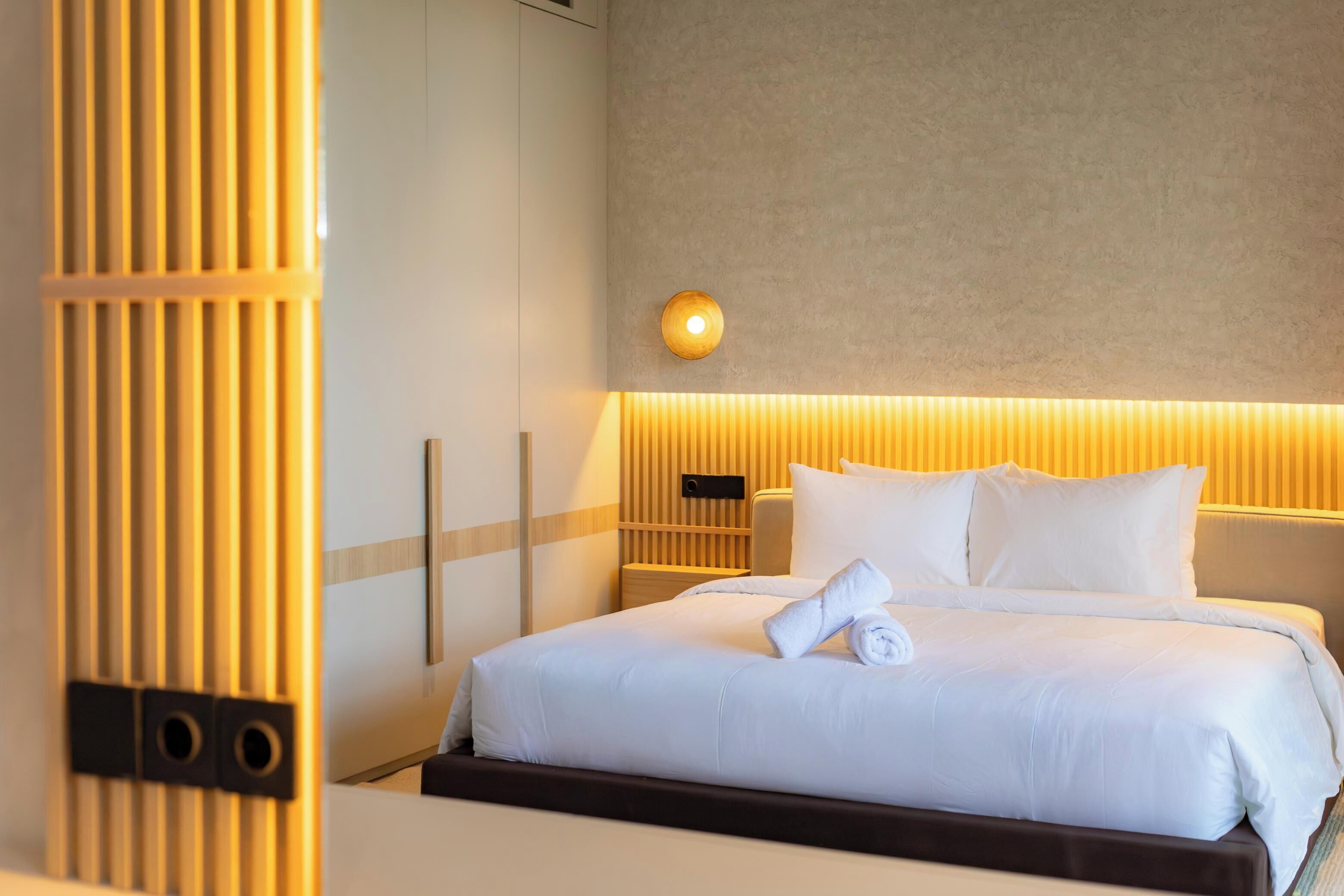 Quarto de hotel com uma cama, mesa de cabeceira, luminária na parede e um painel de madeira vertical.