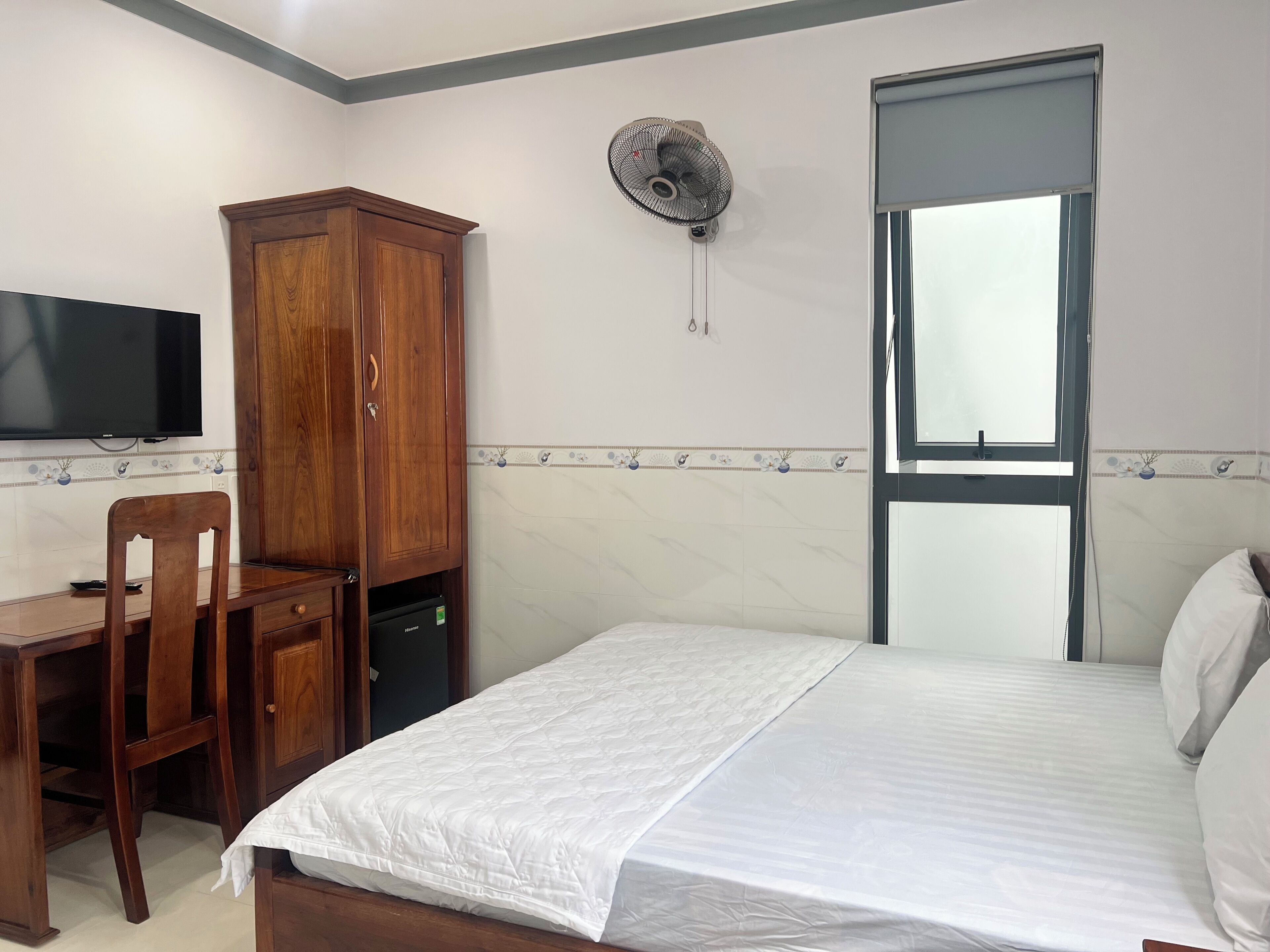 Quarto com cama, guarda-roupa de madeira, escrivaninha com cadeira, televisão e janela.