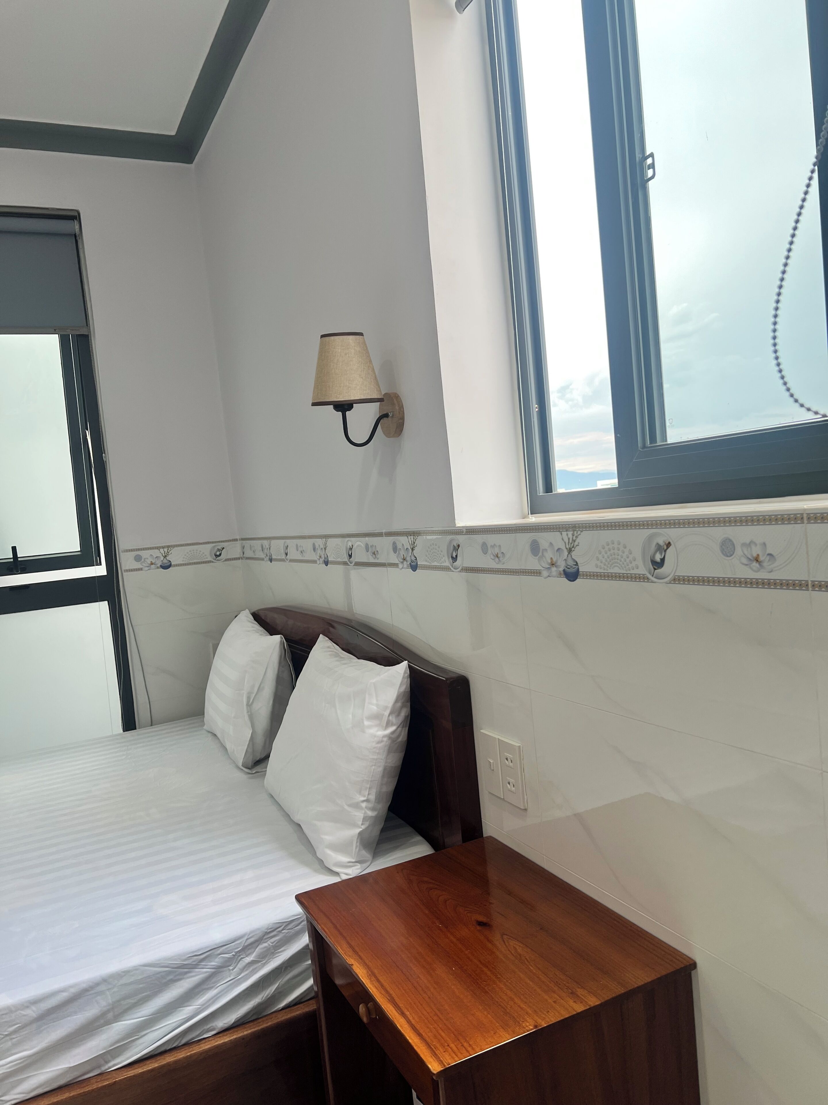 Quarto de hotel com cama, mesa de cabeceira, abajur, janela e parede com moldura decorativa.