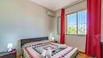 Um quarto com cama, mesa de cabeceira, luminária, janela com cortinas vermelhas e aparelho de ar condicionado.