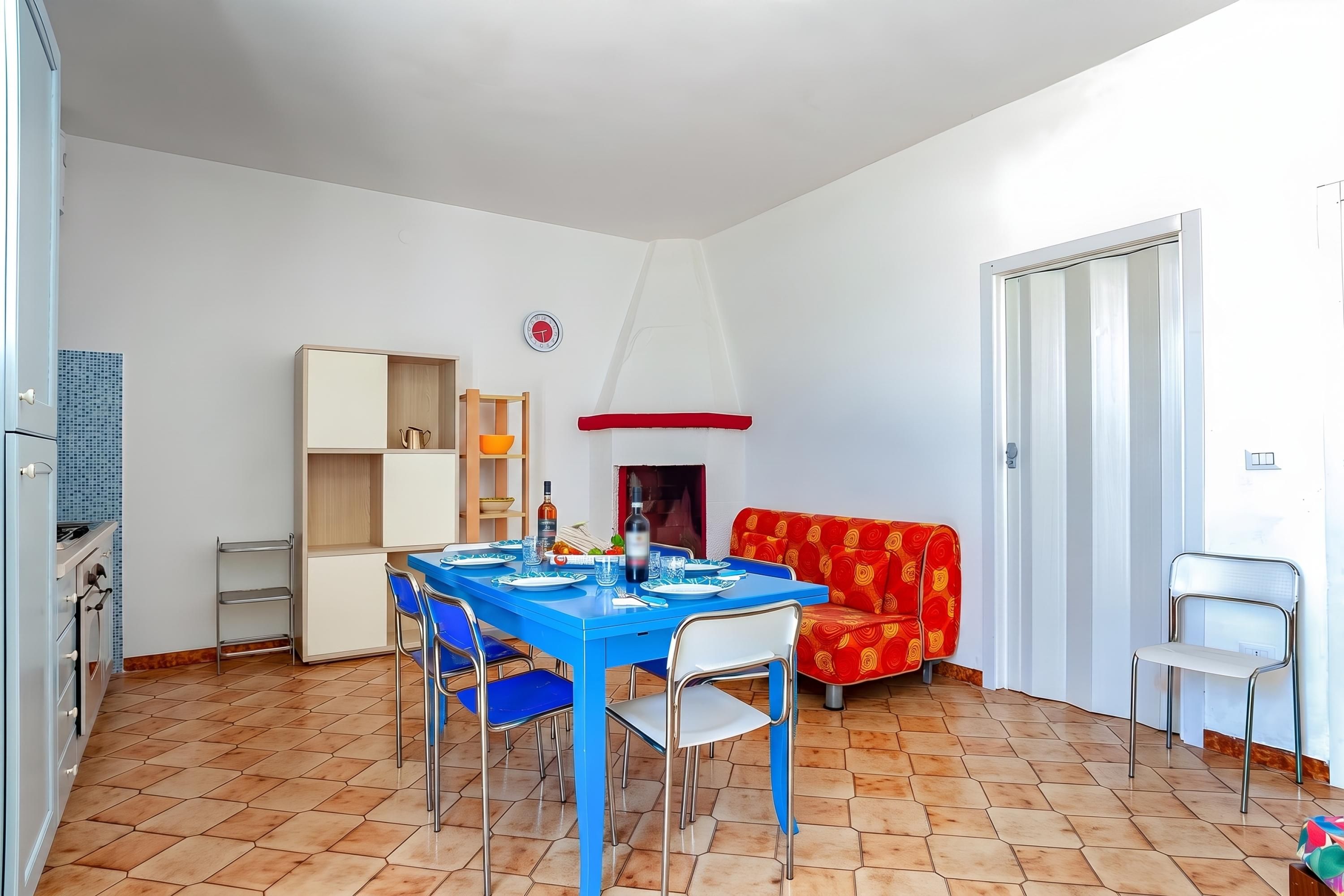 Uma área de refeições com mesa azul, cadeiras e sofá vermelho. Há também uma cozinha com armários e fogão.