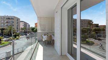Uma varanda com mesa e cadeiras, guarda-corpo de vidro e vista para a rua e edifícios.