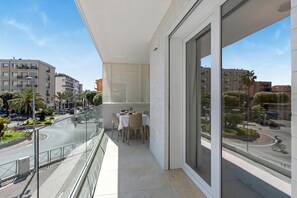 Uma varanda com mesa e cadeiras, guarda-corpo de vidro e vista para a rua e edifícios.