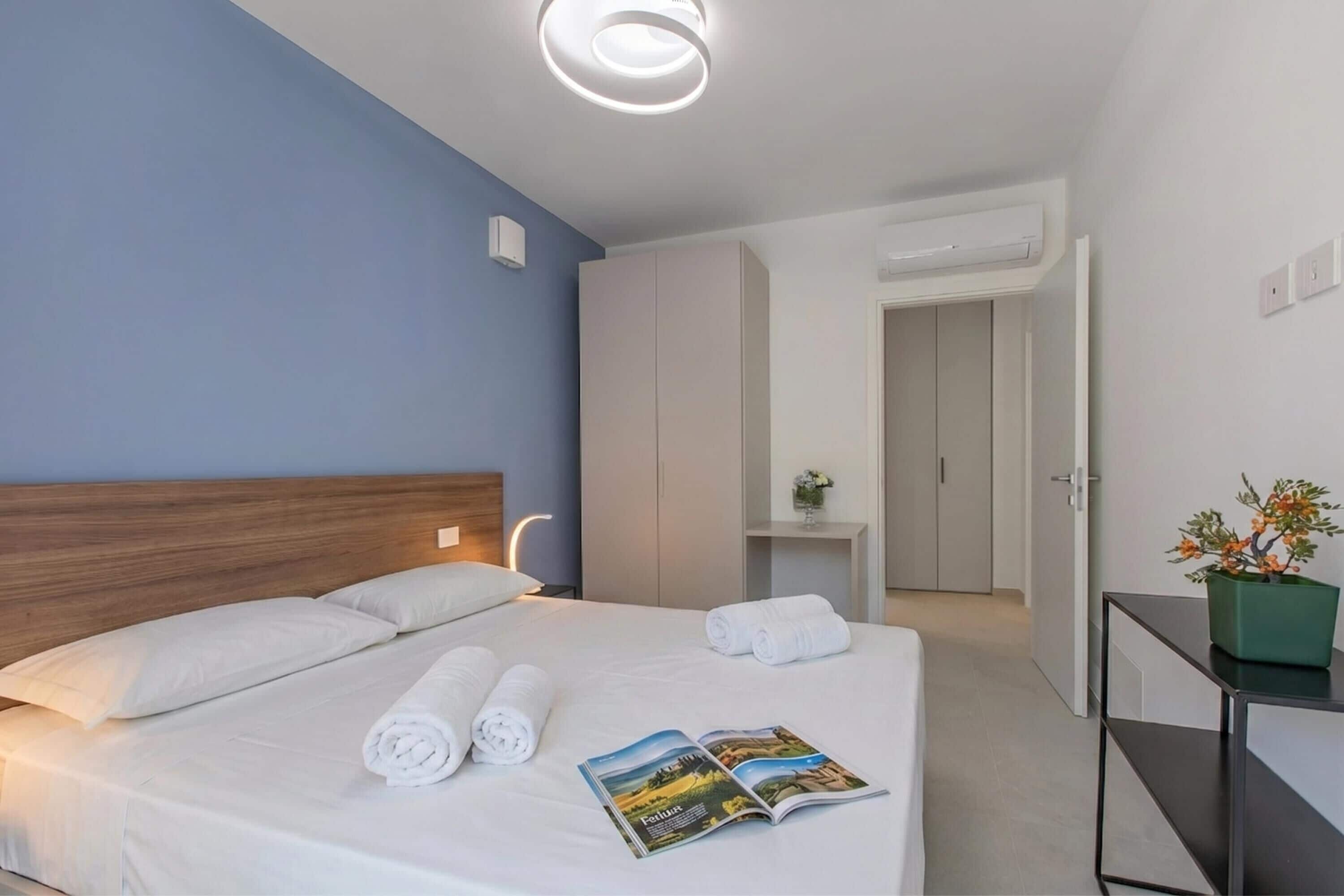 Quarto de hotel com uma cama, mesa de cabeceira com uma revista, planta em vaso e guarda-roupa.