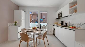 Cozinha moderna com armários brancos, área de jantar com mesa e cadeiras redondas, e vista para prédios através de uma porta de vidro.