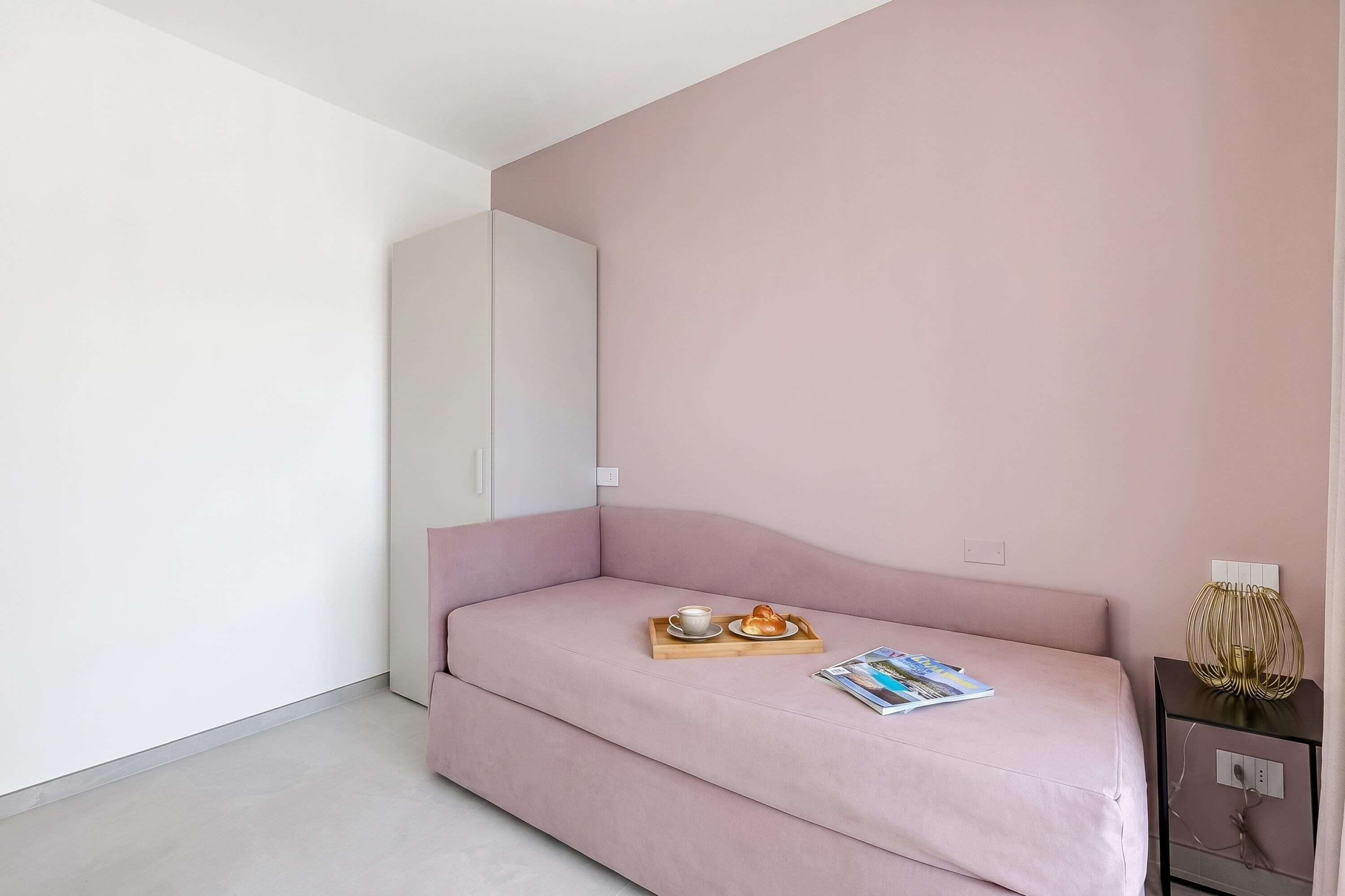 Quarto minimalista com um sofá rosa, uma mesinha com uma chávena e um pastel, uma revista e um candeeiro decorativo numa mesa de cabeceira.