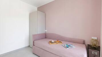 Quarto minimalista com um sofá rosa, uma mesinha com uma chávena e um pastel, uma revista e um candeeiro decorativo numa mesa de cabeceira.