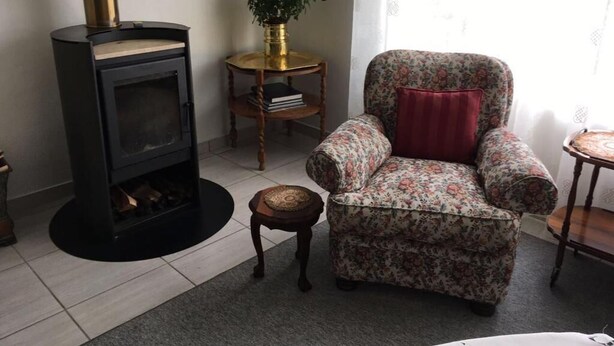 Uma sala de estar com lareira, uma poltrona floral, uma mesa redonda pequena e um vaso com planta.