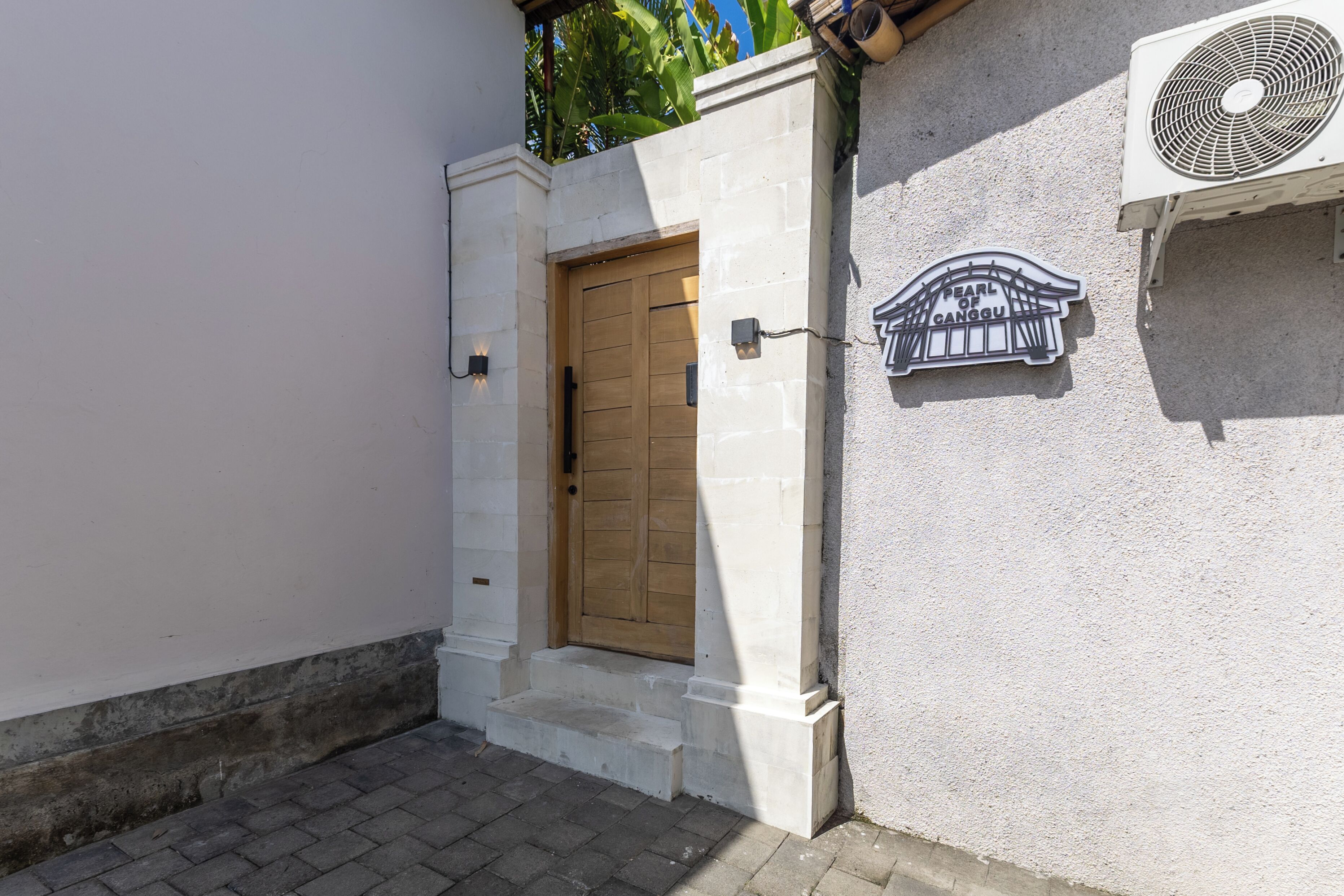 Entrada de um edifício com porta de madeira, ventoinha na parede e uma placa decorativa.