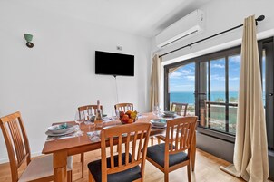 Uma área de refeições com mesa posta para seis, uma TV de tela plana na parede e uma grande janela com vista para o mar.
