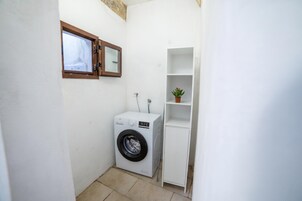 Uma lavanderia compacta com máquina de lavar, uma prateleira pequena com um vaso de planta e uma janela com moldura de madeira.