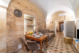 Um interior acolhedor com uma parede de pedra, uma área de jantar com uma mesa posta para dois, um sofá e uma cozinha com geladeira.