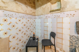 Quarto de canto com ladrilhos estampados, uma mesa com garrafa e copos, e uma cadeira preta.