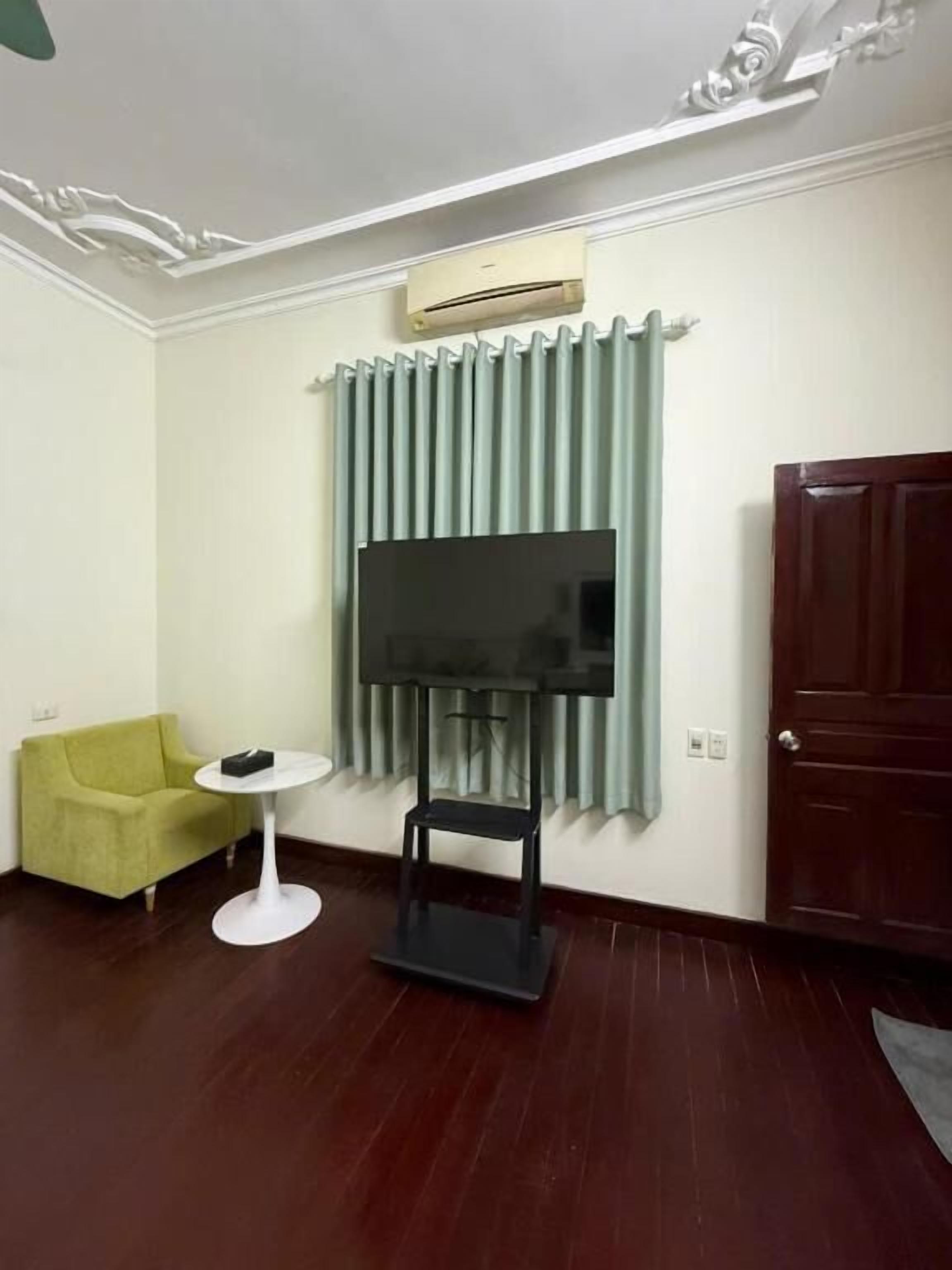 Quarto com televisão de tela plana fixada na parede, sofá verde, mesa redonda pequena e porta de madeira escura.