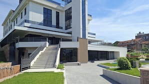 Um edifício moderno de vários andares com uma torre vertical imponente, grandes janelas e uma escadaria que leva à entrada.