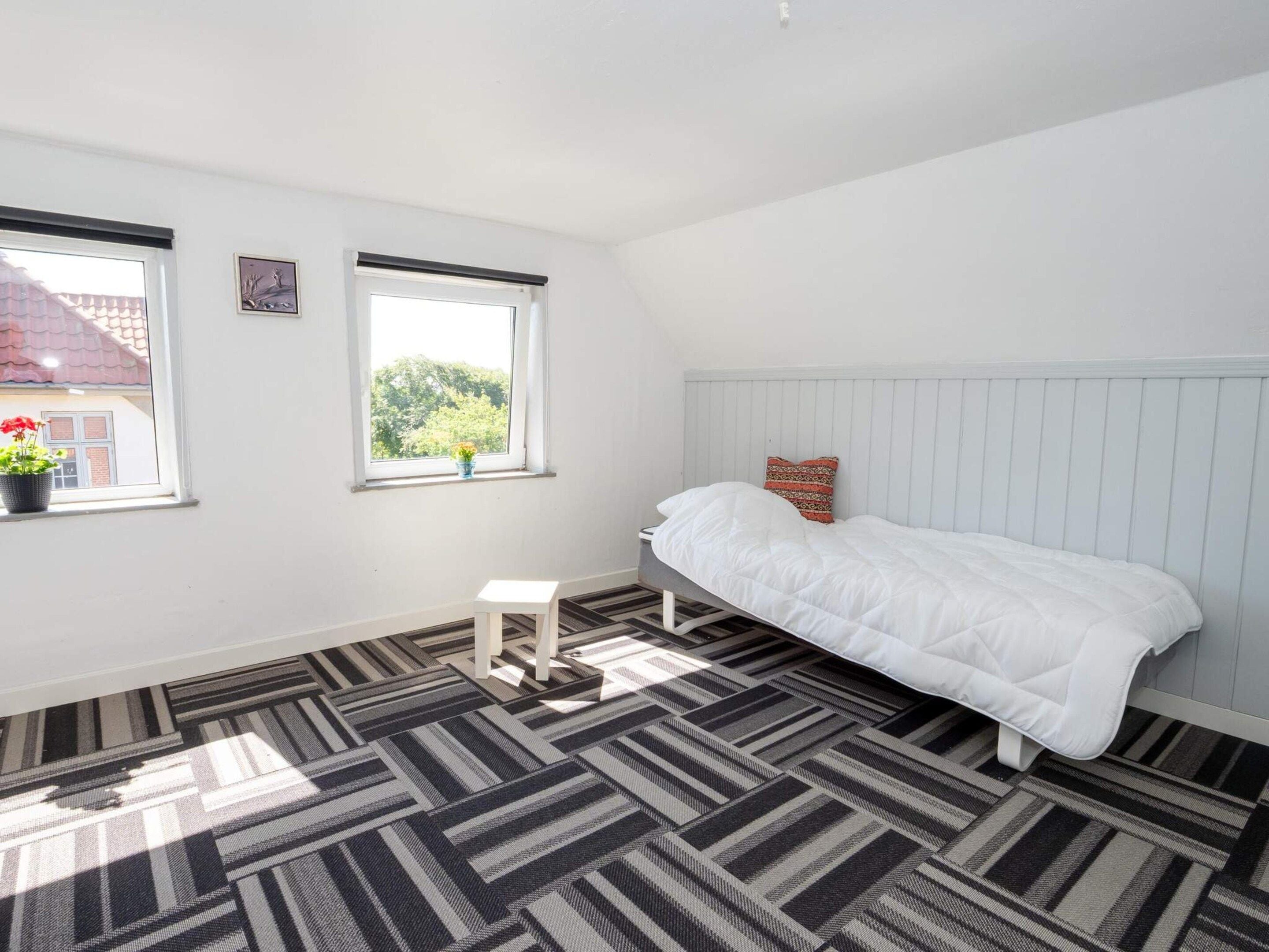 Quarto com cama, duas janelas, uma mesinha de cabeceira e tapete estampado.