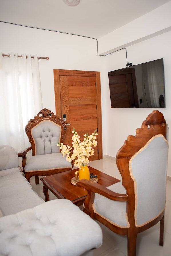 Uma sala de estar com sofá, duas poltronas, uma mesa de centro, uma porta de madeira e uma TV de tela plana na parede.