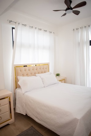 Quarto com cama, mesa de cabeceira, ventoinha no teto e janela com cortinas.