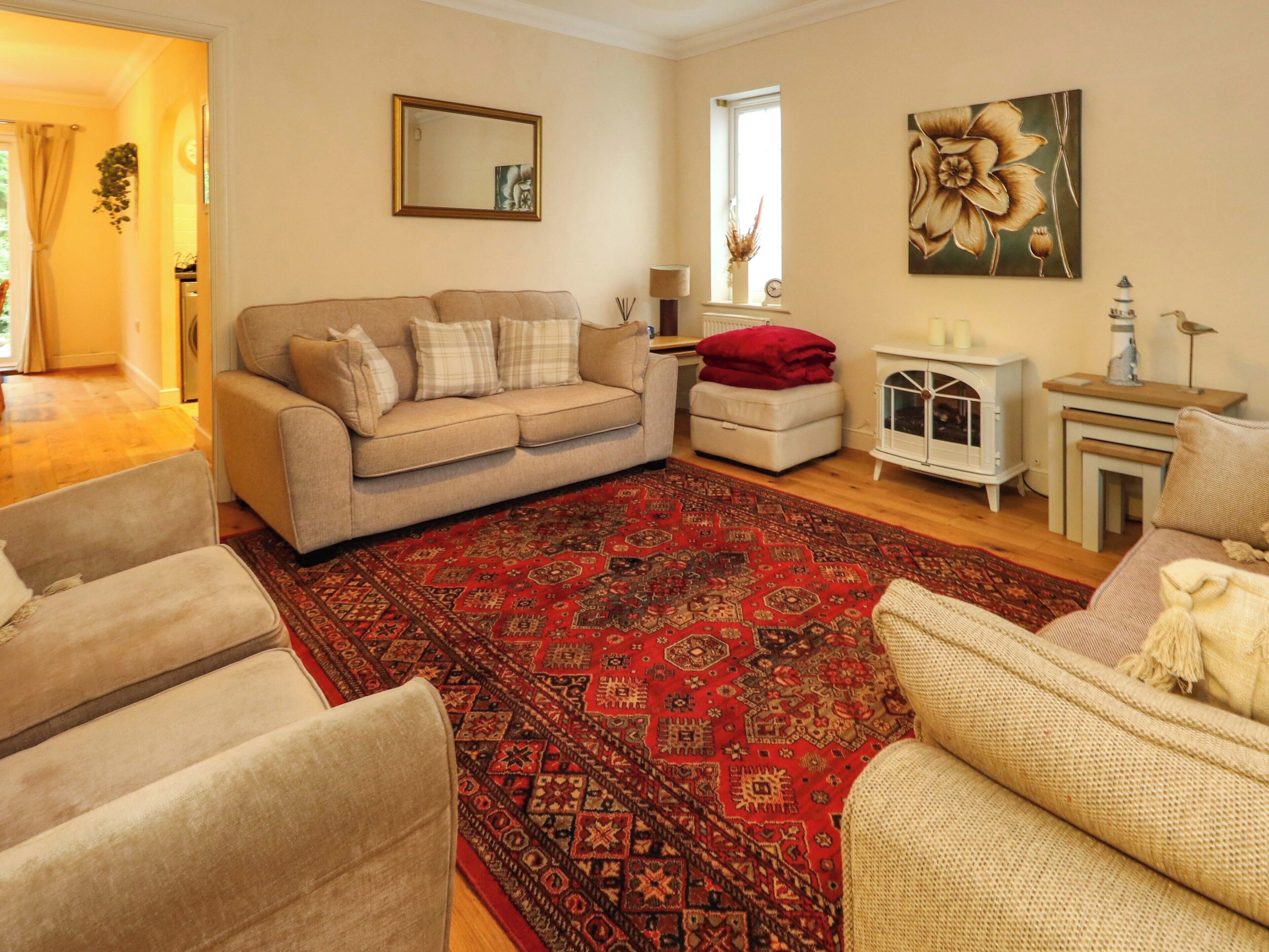 Uma sala de estar com um sofá bege, um tapete estampado, uma mesinha lateral branca e um quadro na parede.