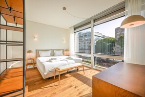 Quarto de hotel moderno com uma cama grande, um banco de madeira, uma escrivaninha e vista da cidade através de amplas janelas.