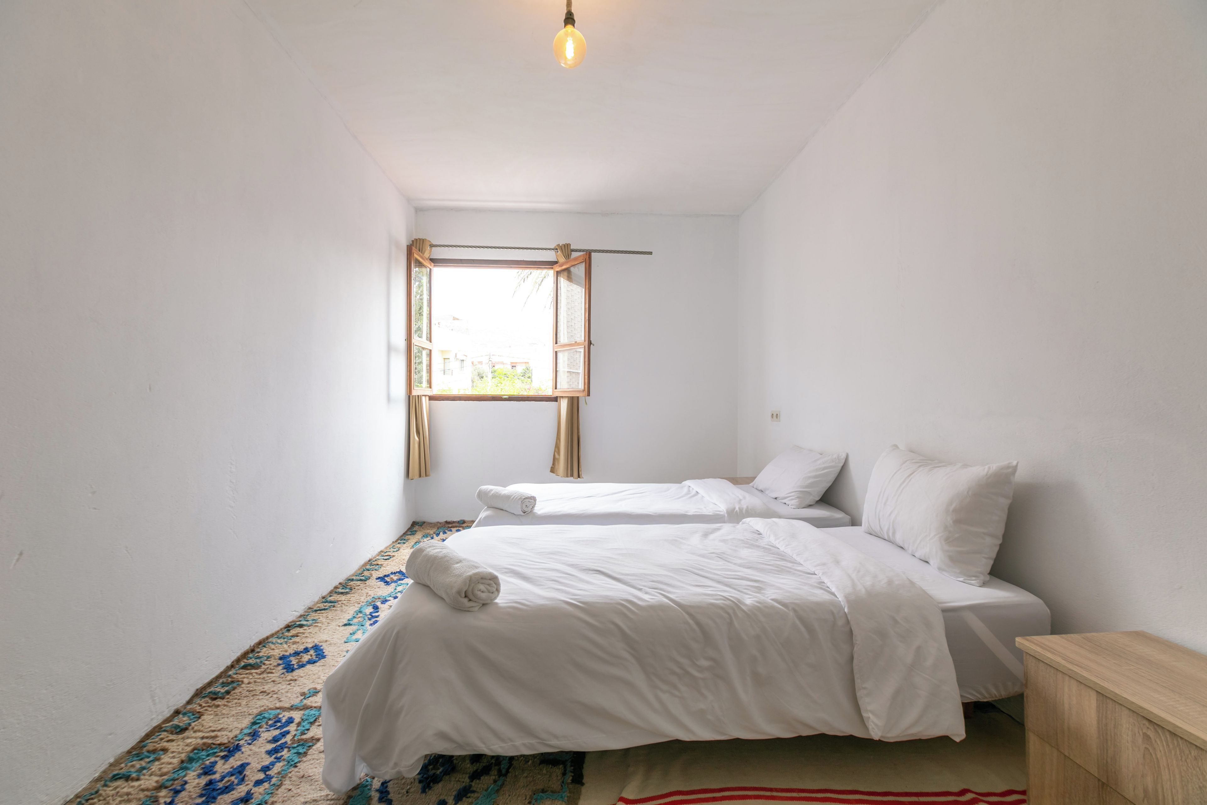 Quarto com duas camas, janela com cortinas, lâmpada pendente e tapete estampado.