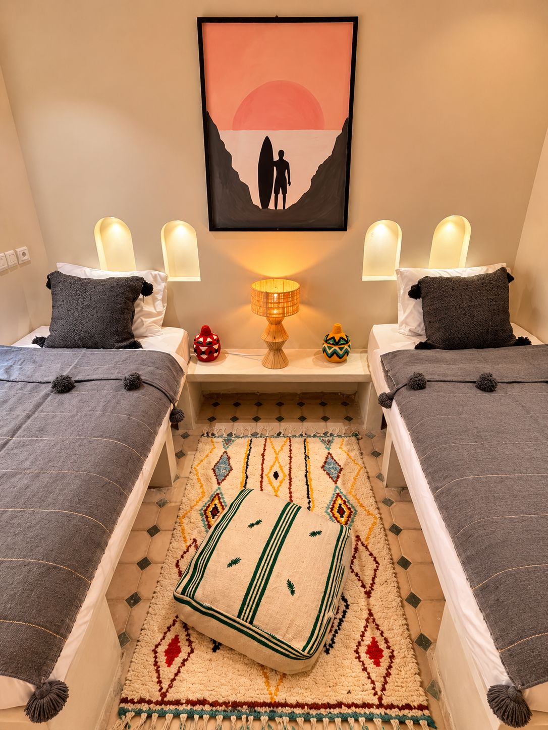 Quarto de hotel com duas camas, um quadro fixado na parede, abajures, almofadas decorativas, um tapete estampado e uma pequena mesa com luminária.