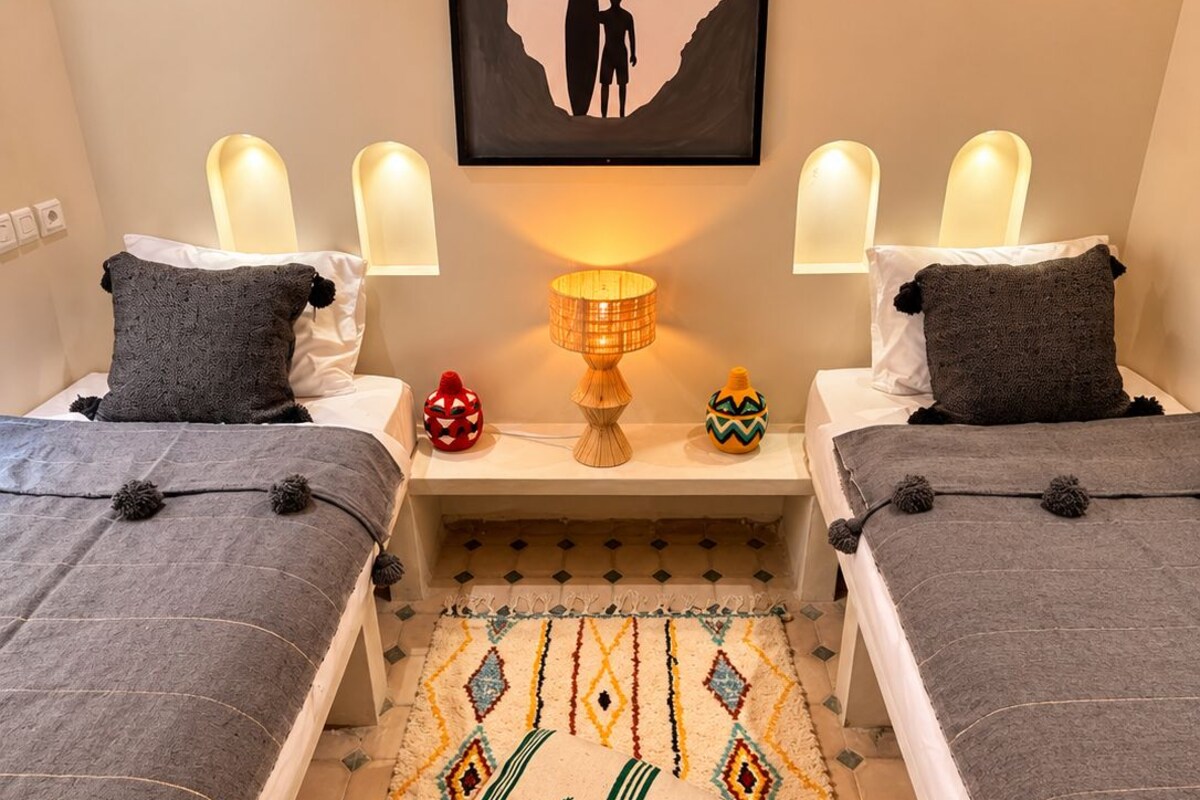 Quarto de hotel com duas camas, um quadro fixado na parede, abajures, almofadas decorativas, um tapete estampado e uma pequena mesa com luminária.
