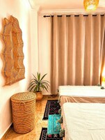 Quarto com cama, cortinas, tapeçaria na parede, planta em vaso, banquinho de vime e tapete estampado.