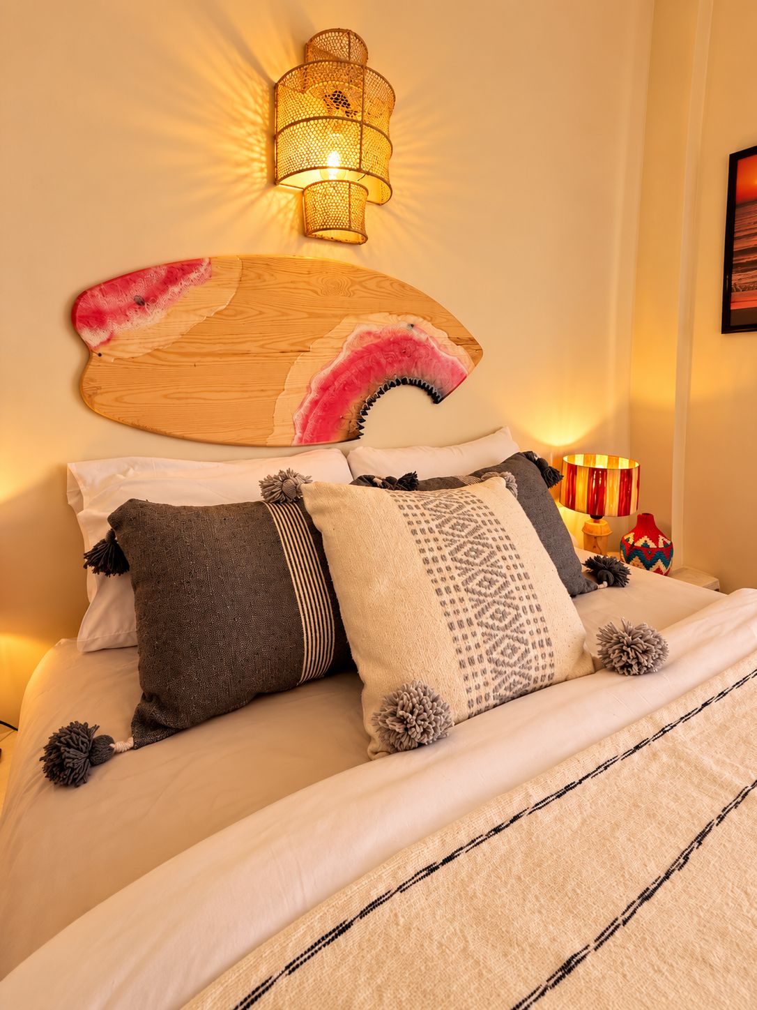 Uma cama com várias almofadas, luminária fixada na parede e um objeto decorativo de madeira sobre a cama.