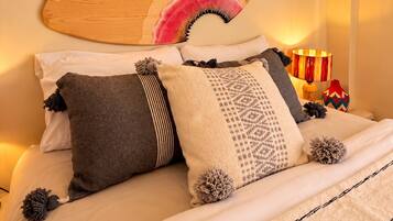 Uma cama com várias almofadas, luminária fixada na parede e um objeto decorativo de madeira sobre a cama.