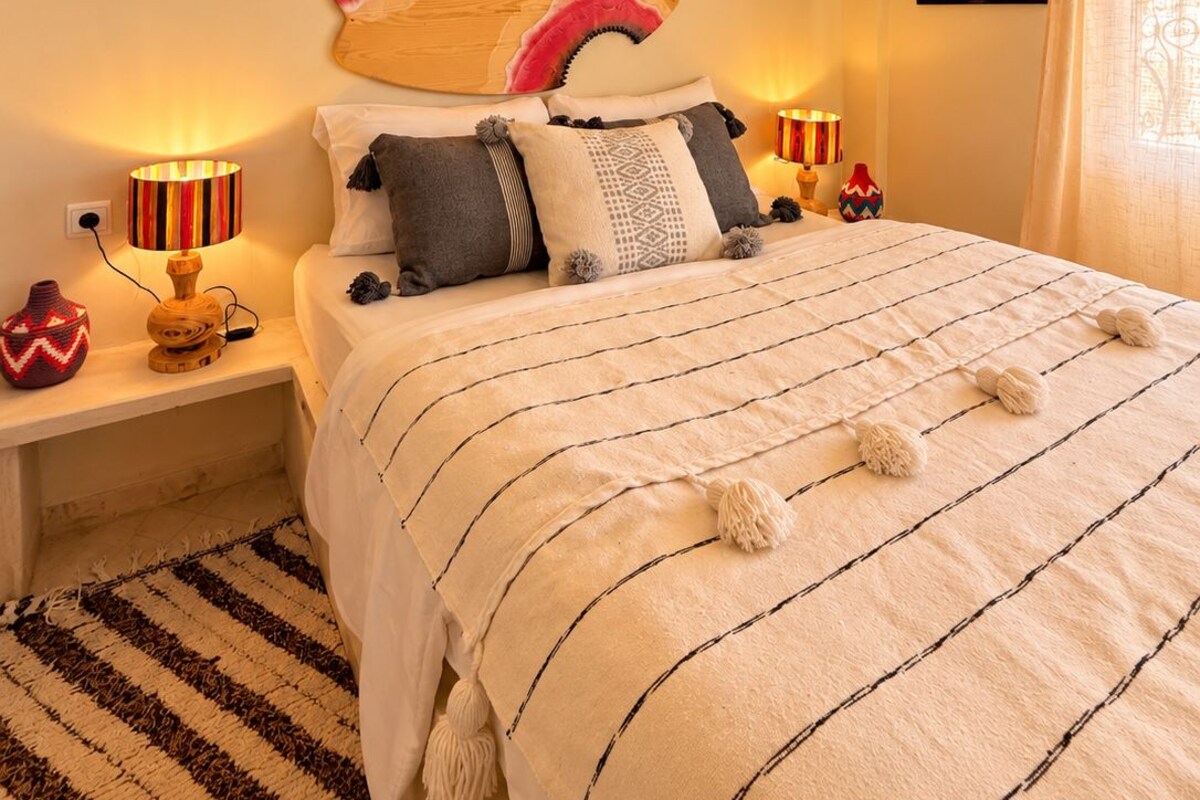 Quarto com cama grande, duas luminárias de cabeceira, luminária na parede, um quadro e janela com cortinas.