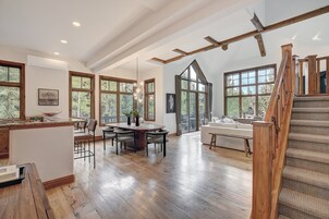 Uma sala de estar ampla com escada de madeira, janelas grandes e área de jantar com mesa e cadeiras.