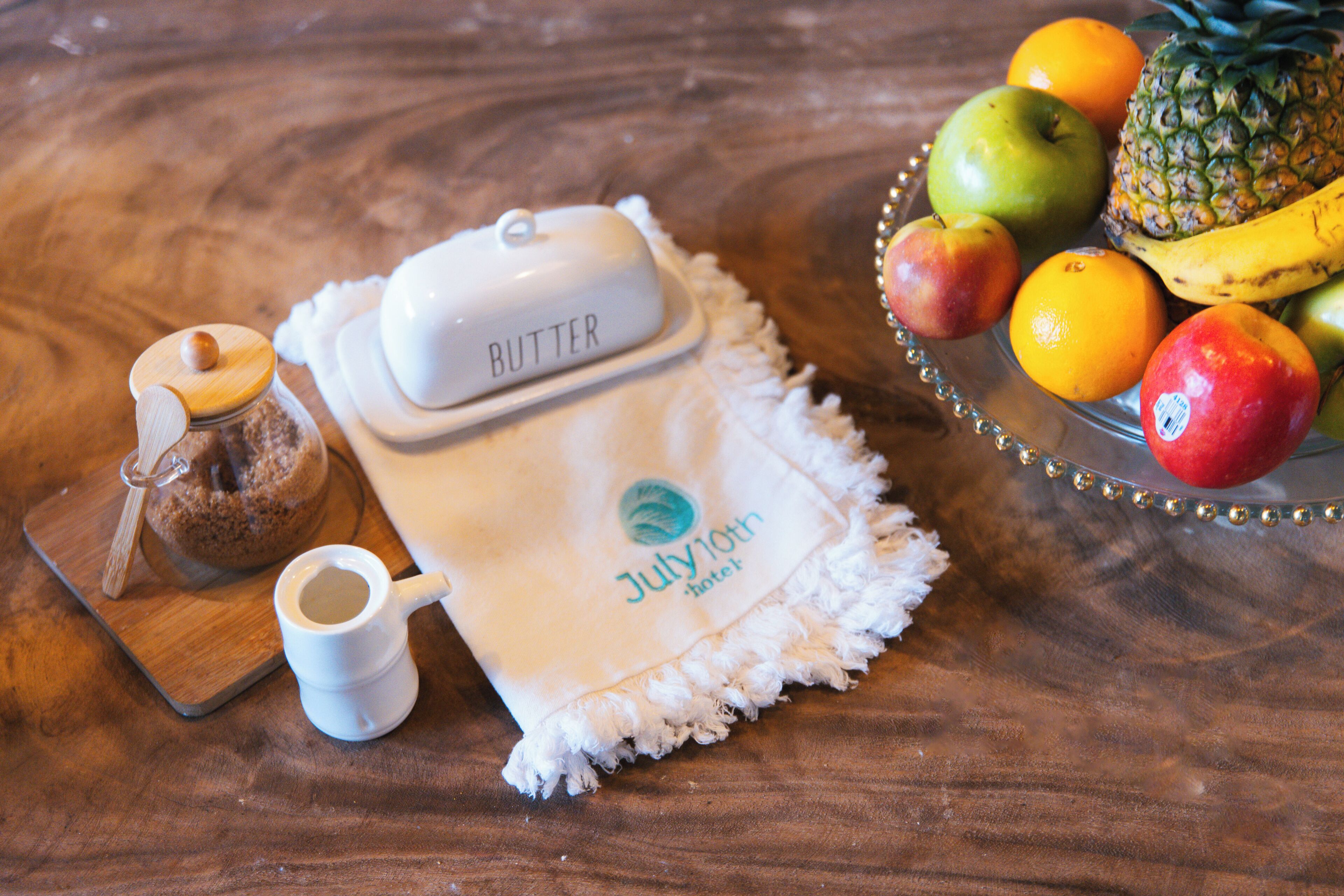 Uma mesa de madeira com um pote de geleia, uma xícara branca pequena, um recipiente para manteiga e um prato com frutas.