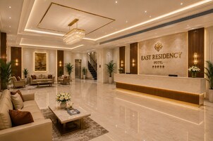 Um saguão de hotel com balcão de recepção, sofás, um lustre e uma escadaria.