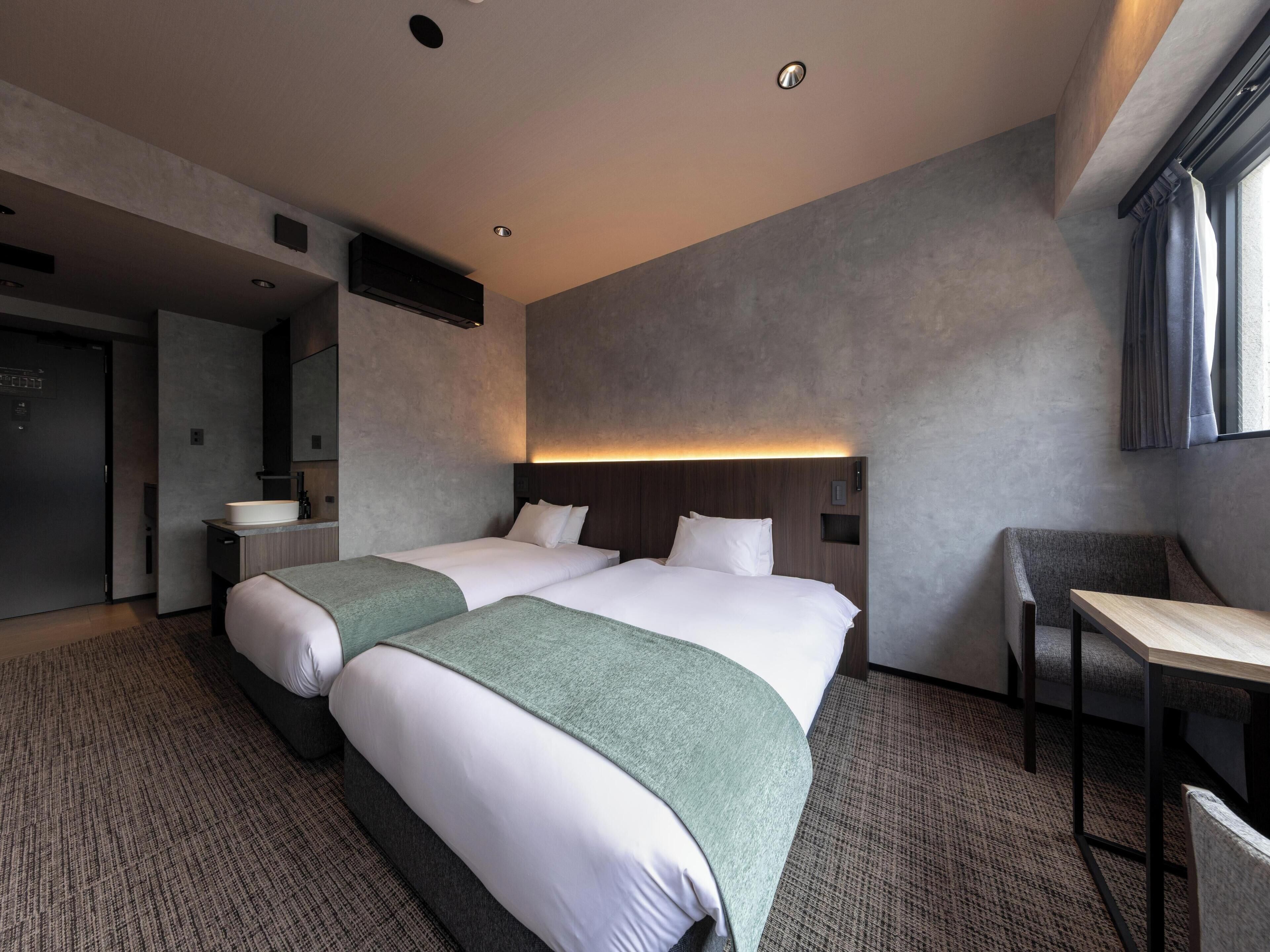 Quarto de hotel com duas camas, uma escrivaninha, uma cadeira, banheiro e uma janela grande.