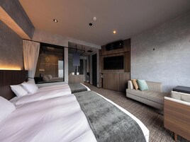 Quarto de hotel moderno com uma cama grande, um sofá, uma TV fixada na parede e um banheiro visível através de uma porta de vidro.