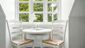 Um quarto branco com duas cadeiras brancas, uma mesa redonda pequena e uma janela com vegetação lá fora.
