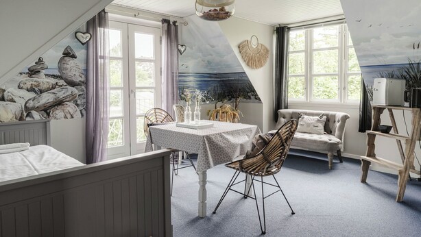 Um quarto com teto inclinado, uma mesa de jantar com cadeiras, um sofá, um mural decorativo na parede e uma janela grande.