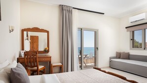 Quarto com cama, escrivaninha com cadeira, sofá, espelho grande e porta de vidro deslizante para uma varanda com vista para o mar.