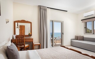 Quarto com cama, escrivaninha com cadeira, sofá, espelho grande e porta de vidro deslizante para uma varanda com vista para o mar.