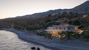 Uma propriedade costeira com praia, uma casa com terraço e montanhas ao fundo.