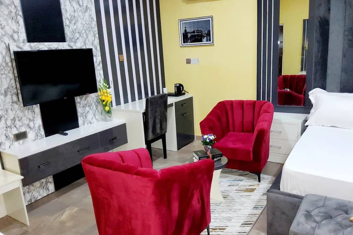 Quarto de hotel com uma cama, duas poltronas vermelhas, uma televisão, uma escrivaninha e um espelho.