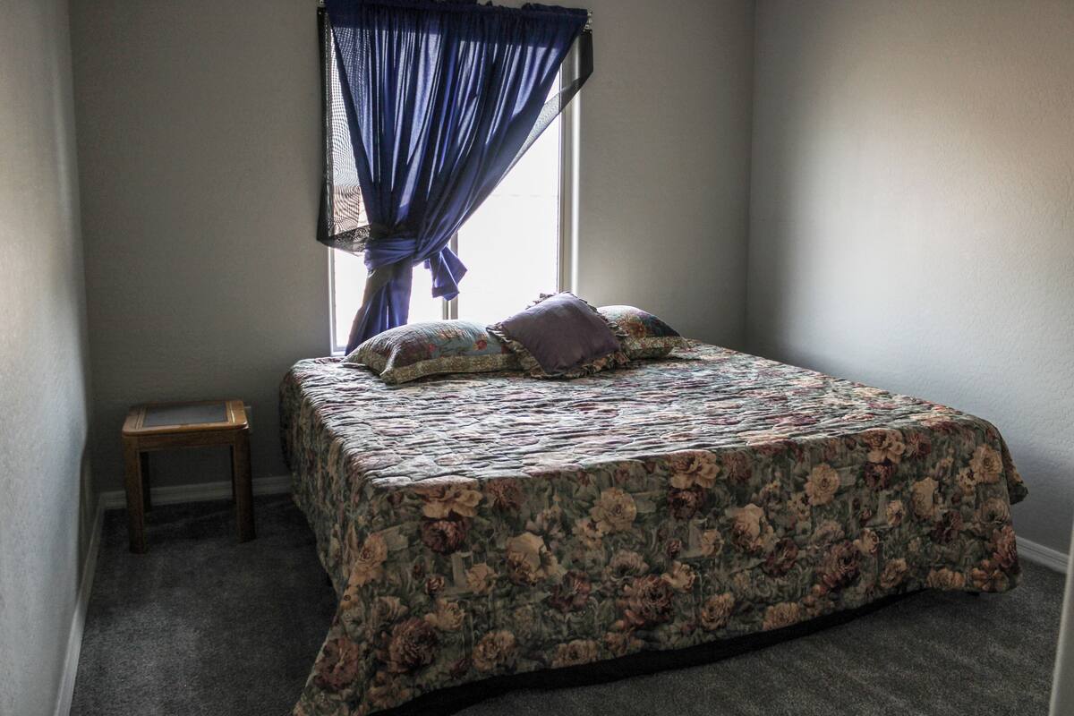 Quarto com cama, um banquinho e janela com cortinas azuis.
