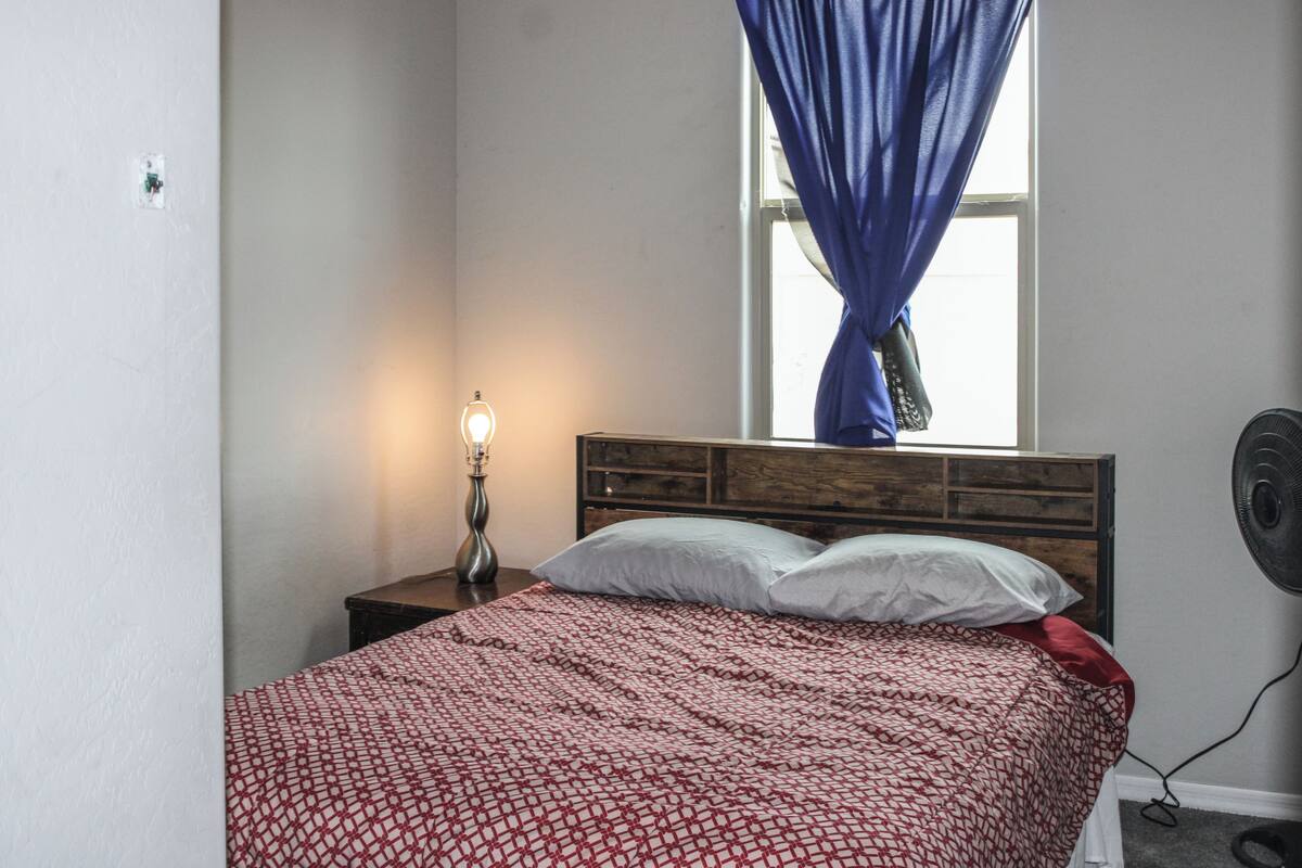 Quarto com cabeceira de madeira, mesa de cabeceira com abajur, janela com cortinas azuis e cama com colcha estampada.
