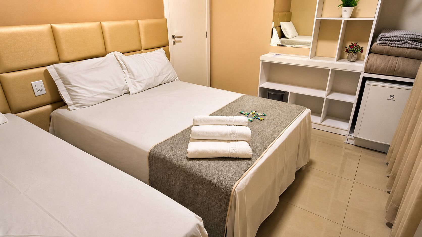 Quarto de hotel com duas camas, uma escrivaninha, um espelho, uma prateleira com uma planta e um armário.
