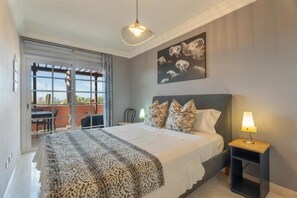 Quarto com cama, mesa de cabeceira, luminária, iluminação no teto, uma obra de arte na parede e vista para área externa por uma porta de correr.